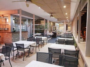 Espaço de refeições interno com mesas, cadeiras, ventilador de teto e vista para a rua.