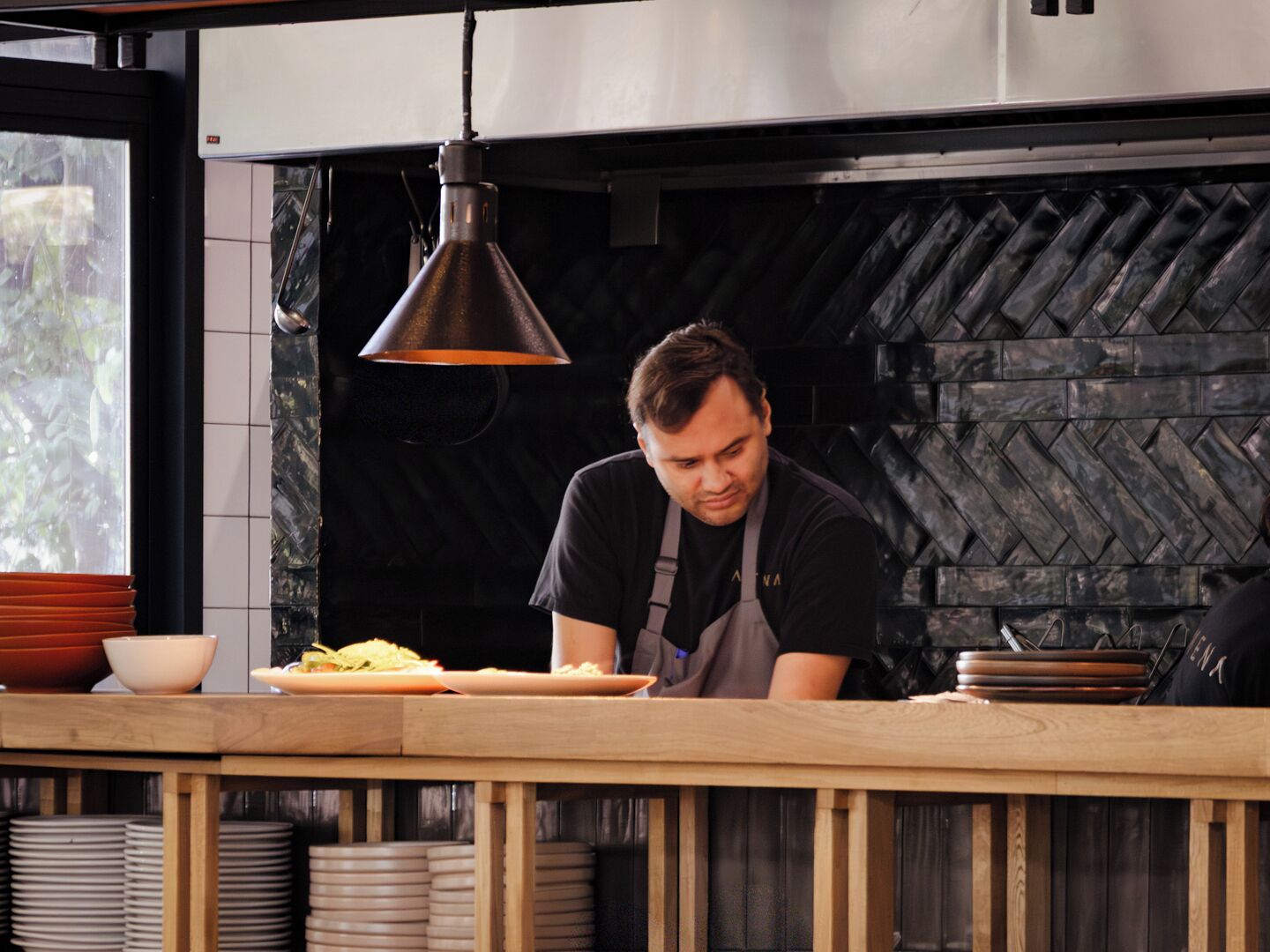 Um chef em um balcão, com uma cozinha moderna ao fundo, pratos empilhados e uma luminária pendente.