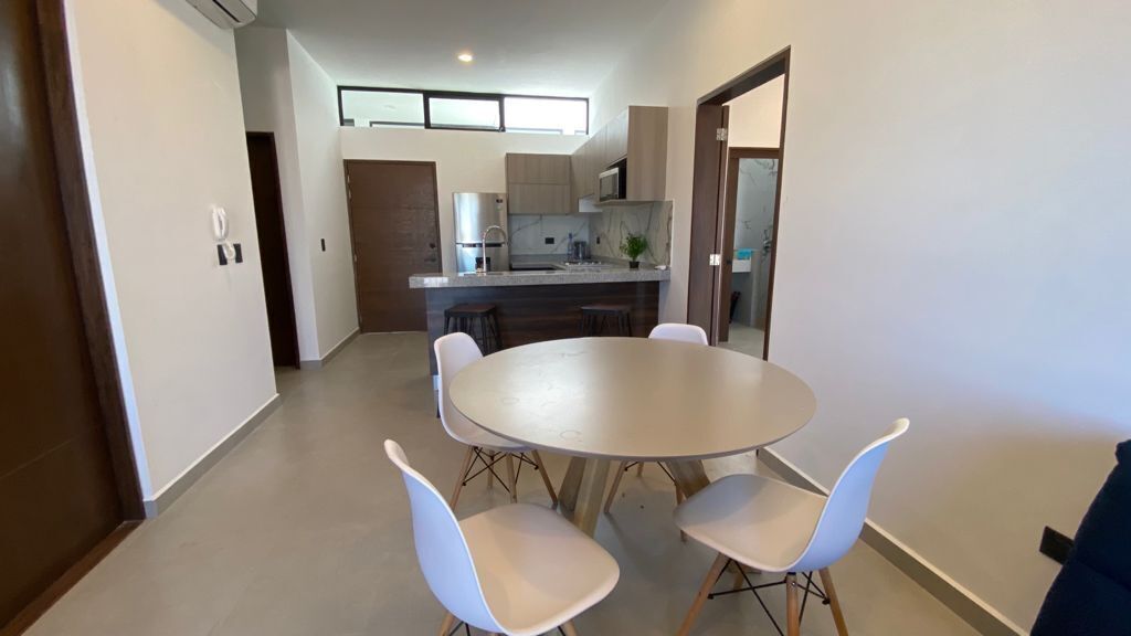 Uma cozinha moderna com uma ilha central, uma sala de jantar com mesa redonda e cadeiras brancas, e um corredor que leva a uma porta.