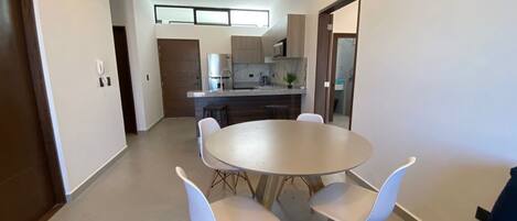 Uma cozinha moderna com uma ilha central, uma sala de jantar com mesa redonda e cadeiras brancas, e um corredor que leva a uma porta.