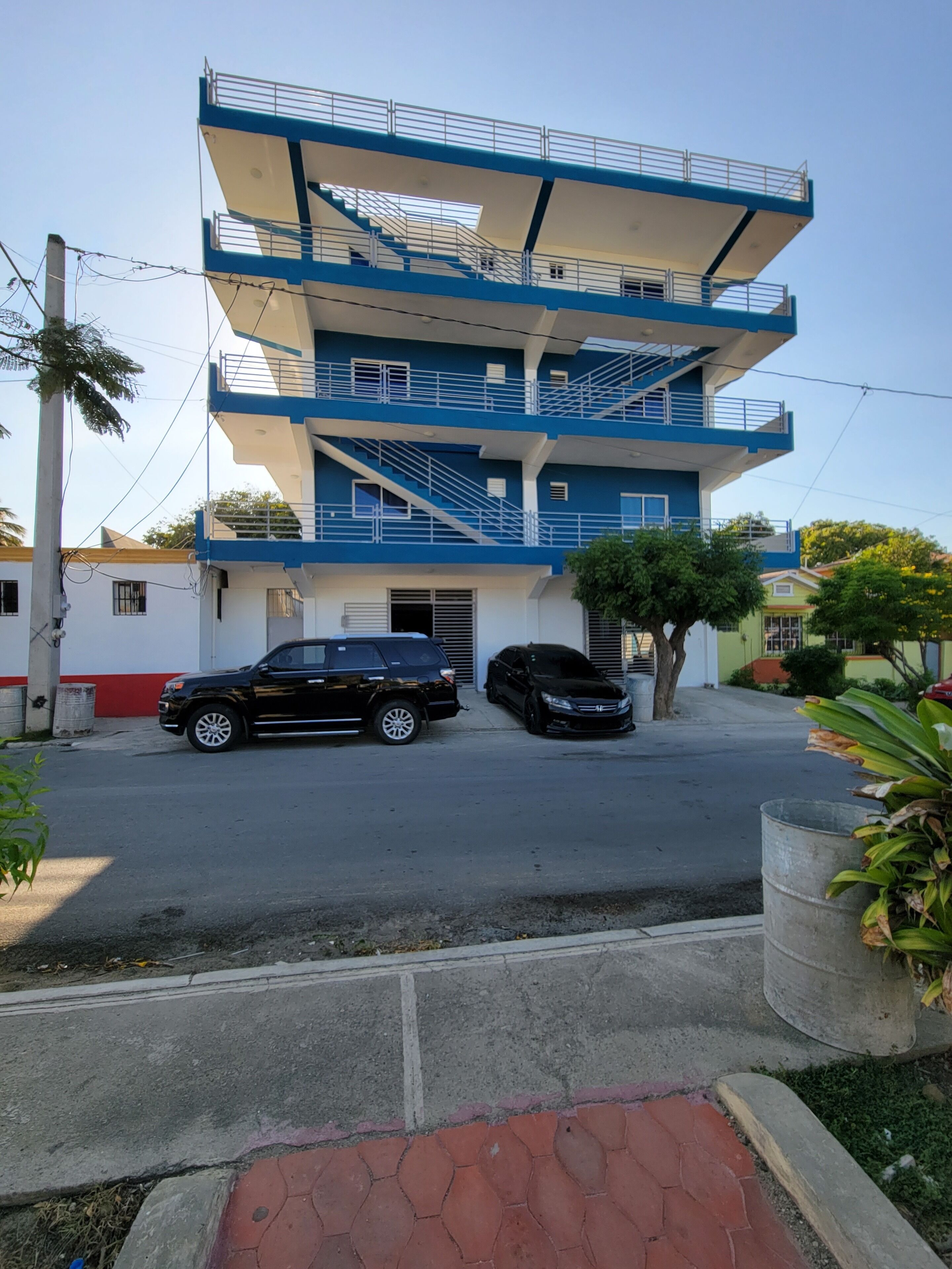Um edifício de vários andares com esquema de cores azul e branco, apresentando uma escadaria e uma varanda. Dois carros estão estacionados na frente.