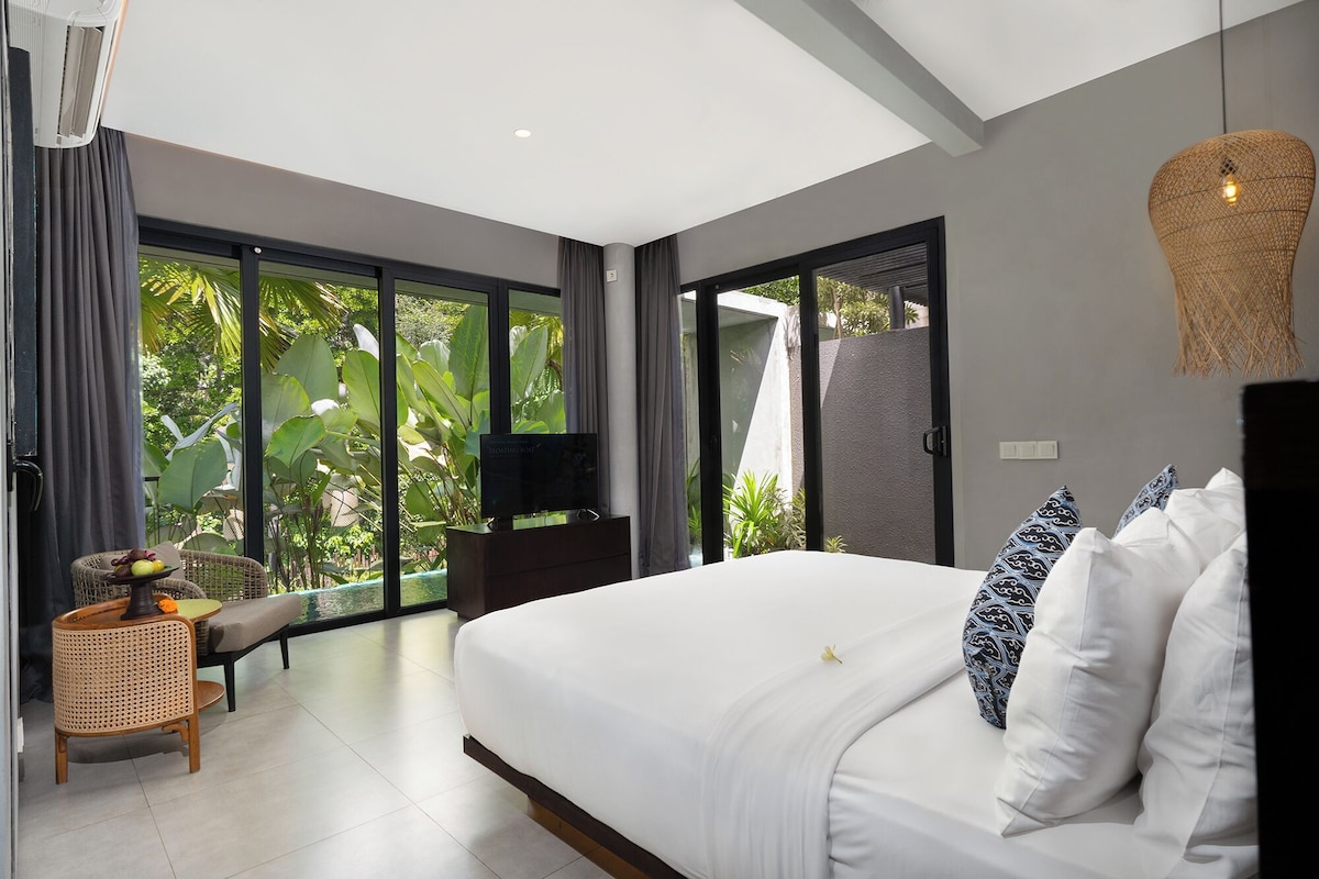 Quarto moderno com uma cama grande, televisão e vista para vegetação exuberante pelas janelas.