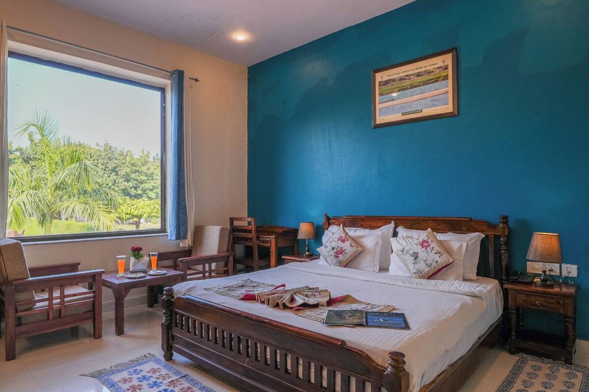 Quarto com uma janela grande, cama com almofadas florais, cabeceira de madeira, mesa de cabeceira com abajur e vista para o verde lá fora.