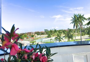 Vista de uma varanda com vista para uma praia, palmeiras, piscina e céu claro.