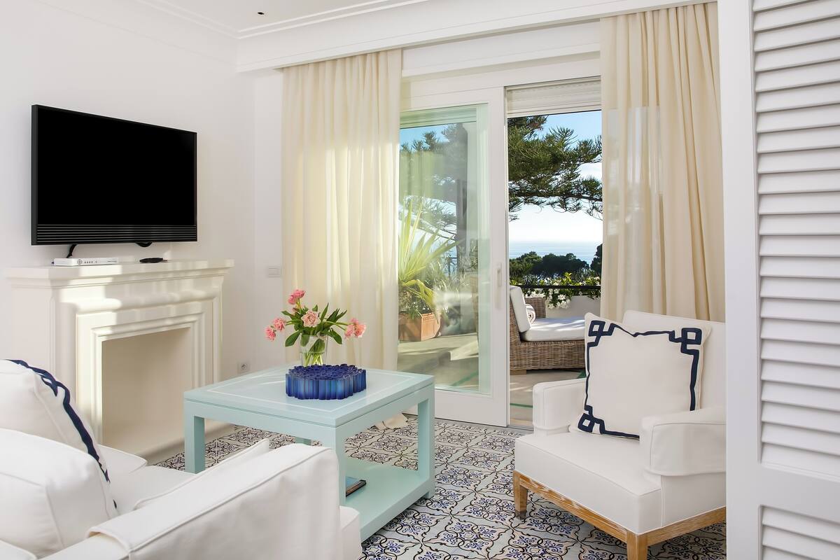 Uma sala de estar com uma TV de tela plana, uma lareira, uma mesa de centro com um vaso de flores e vista para o exterior por meio de grandes portas de vidro.