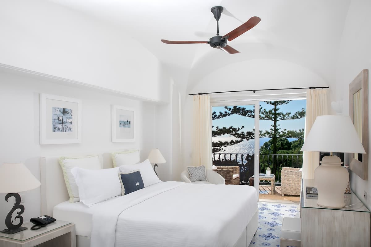 Quarto de hotel com cama, duas mesas de cabeceira com luminárias, ventoinha no teto e varanda com mobiliário.