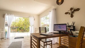 Quarto com escrivaninha de madeira, computador e cadeira, com janelas amplas e vista para o verde lá fora.