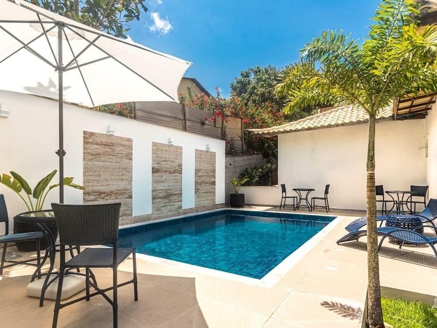 Piscina com espreguiçadeiras e uma mesa, cercada por um pátio ladrilhado e um pequeno jardim.