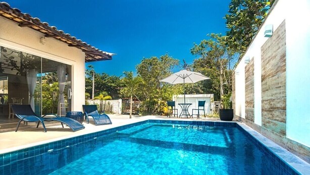 Piscina com espreguiçadeiras e mesa, cercada por um edifício com telhado telhado e área de jardim.