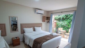 Quarto de hotel com cama, mesa de cabeceira, espelho, ar condicionado e vista para área externa com cadeiras e uma mesa.