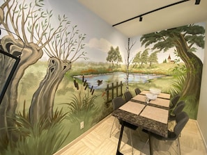 Uma área de refeições com um mural que retrata uma paisagem com árvores, um rio e um edifício ao longe.