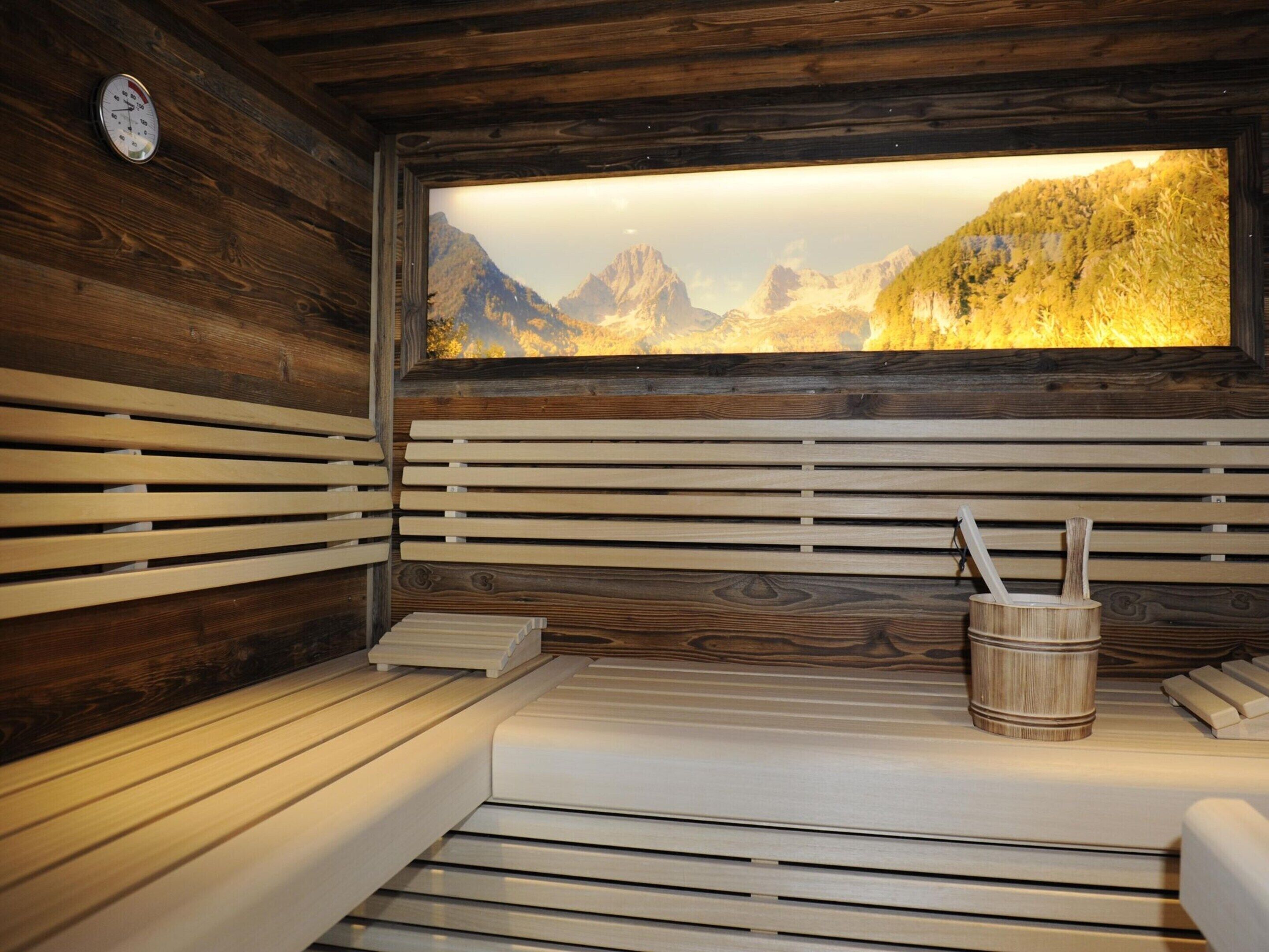 Uma sauna com paredes de madeira, uma janela grande com vista para as montanhas, um balde de madeira e um banco.