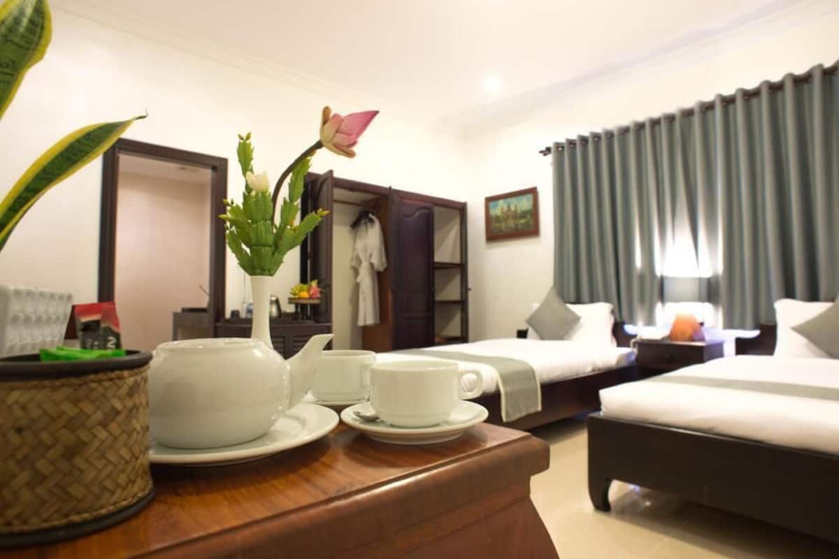 Quarto de hotel com cama, mesa de cabeceira, planta em vaso, quadro e janela com cortinas.