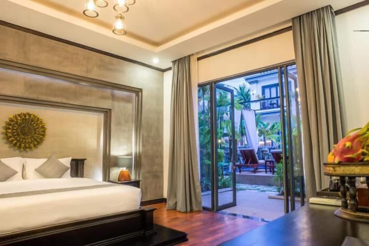 Quarto com cama grande, mesa de cabeceira, luminária, detalhe decorativo na parede e vista para área externa com piscina e espreguiçadeiras.