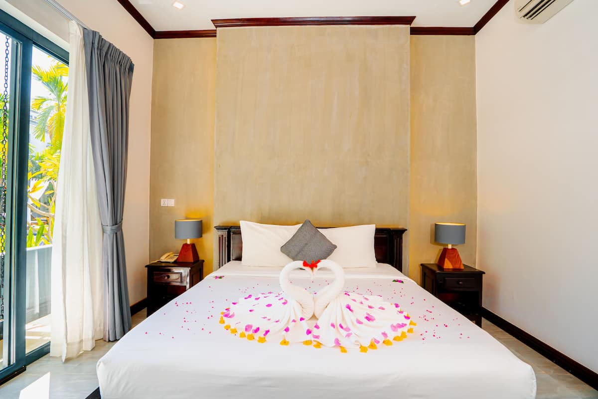 Quarto de hotel com uma cama grande, duas mesas de cabeceira com abajures, uma janela com cortinas e vista para palmeiras.