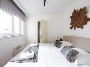 Quarto de hotel com cama, mesa de cabeceira, um objeto decorativo na parede e uma janela com persianas.
