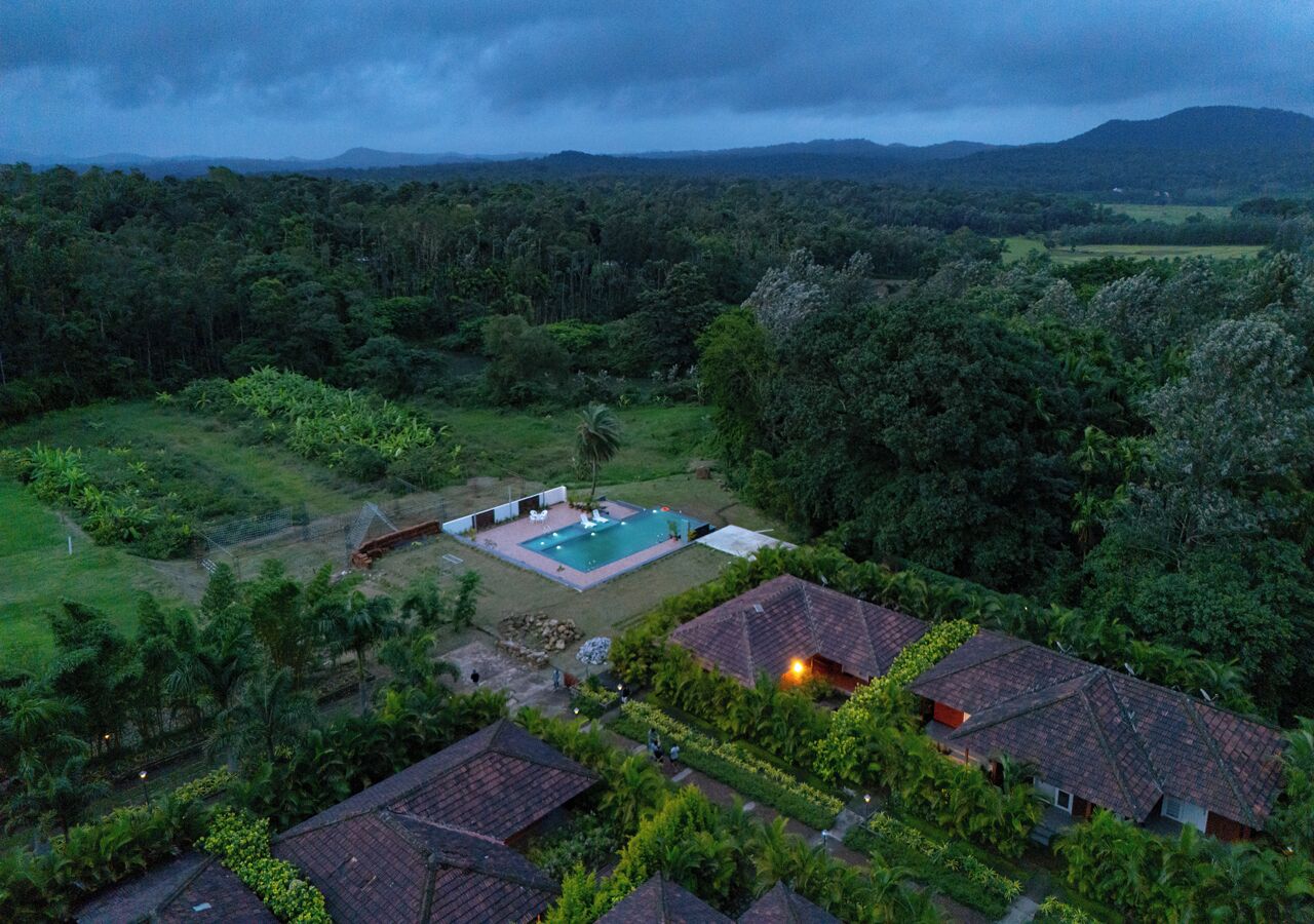 Vista aérea de um resort com piscina, rodeado por vegetação exuberante e colinas ao fundo.