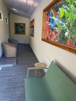Um corredor estreito com piso de madeira, um banco acolchoado verde e uma pequena mesa redonda. Há duas fotos emolduradas na parede, uma representando uma floresta e a outra com flores coloridas.