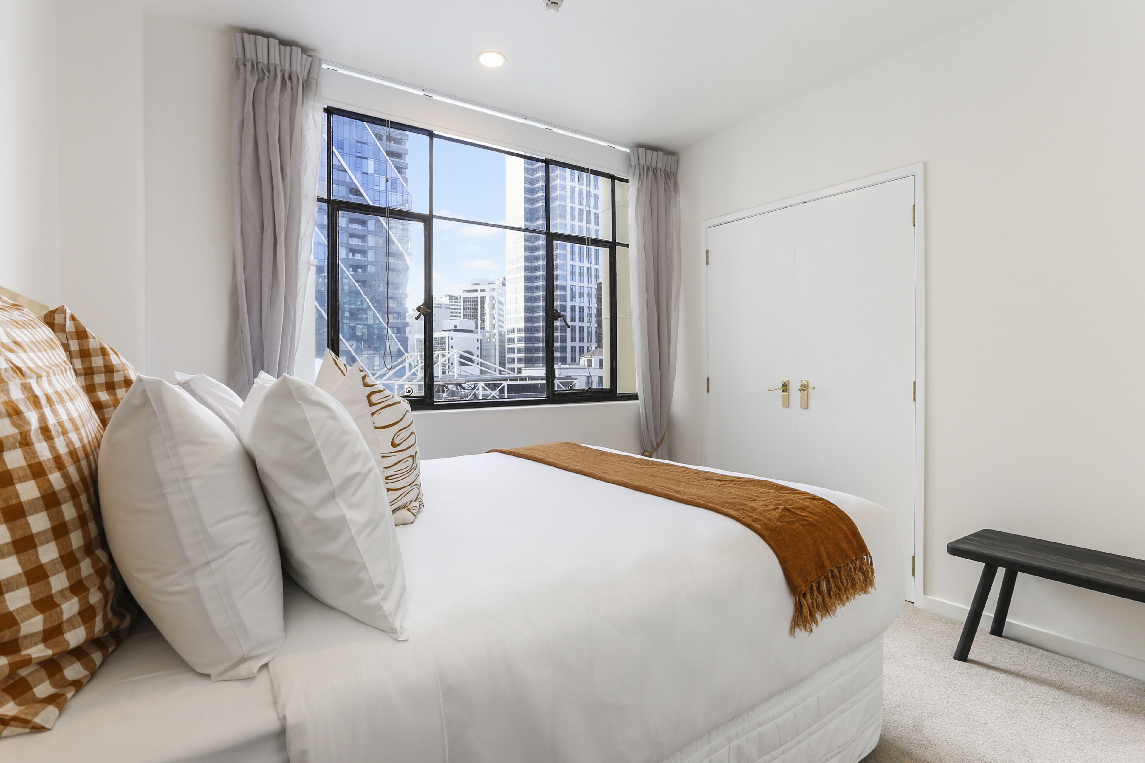 Quarto de hotel com cama grande, janela com vista para a cidade, um banco e uma porta.