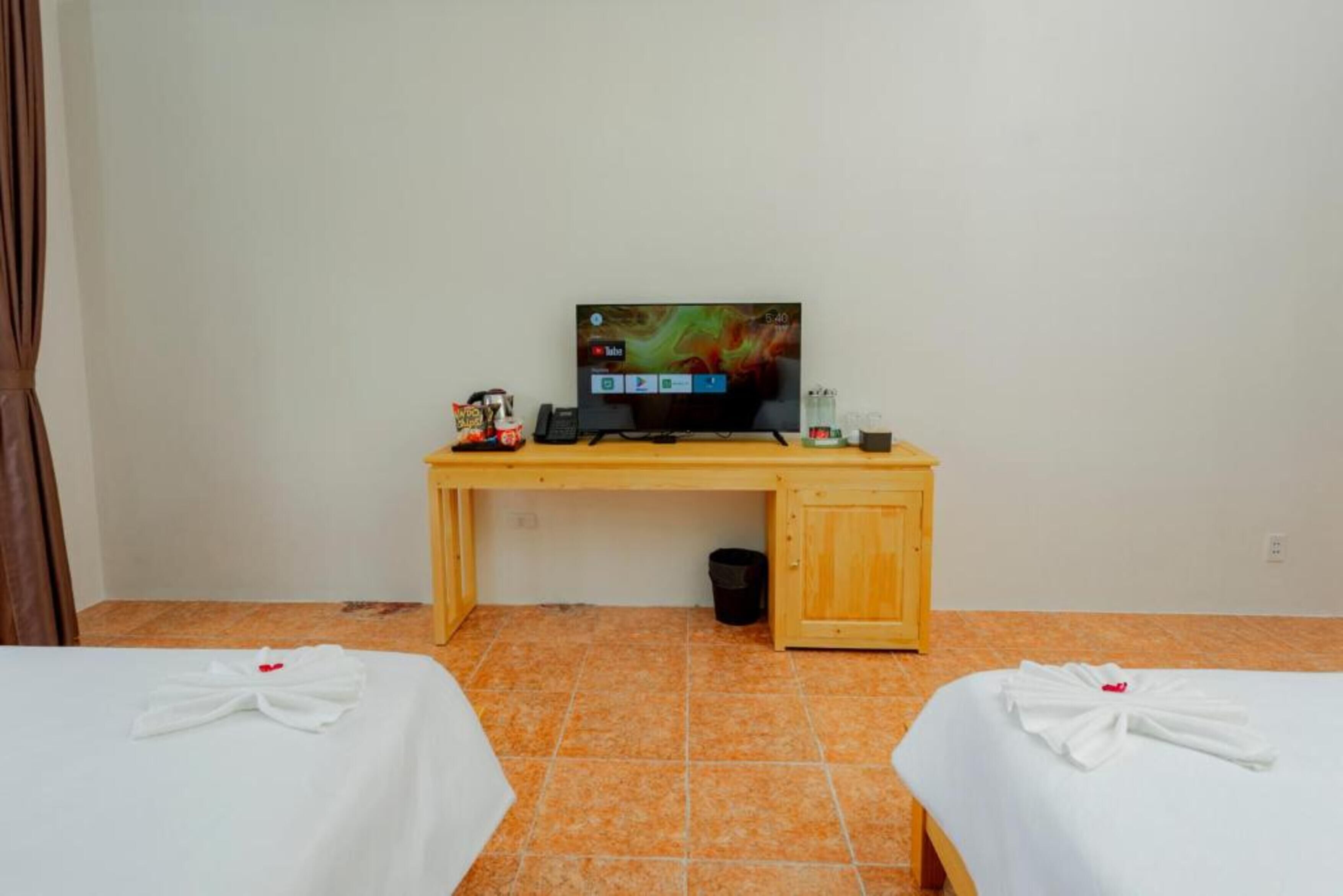 Quarto de hotel com duas camas, uma escrivaninha de madeira com uma TV, um cesto de lixo e uma pequena mesa com objetos.