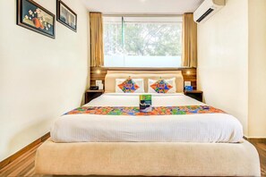 Uma cama bem arrumada com um edredom colorido, uma mesa de cabeceira com um livro e uma janela com cortinas.