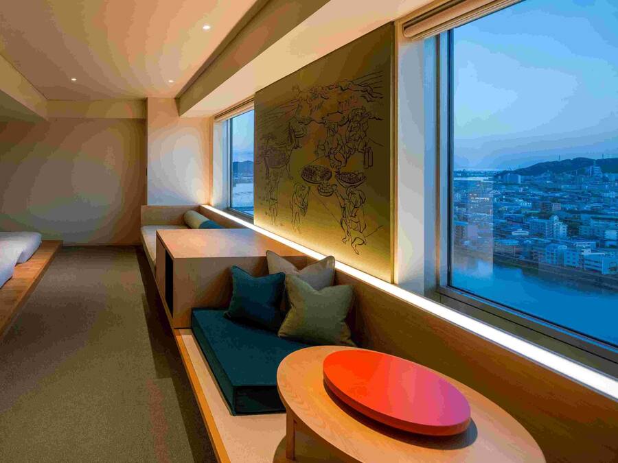 Quarto de hotel moderno com uma grande janela oferecendo vista da cidade, um espaço de estar acolhedor com almofadas e uma mesa vermelha distinta.