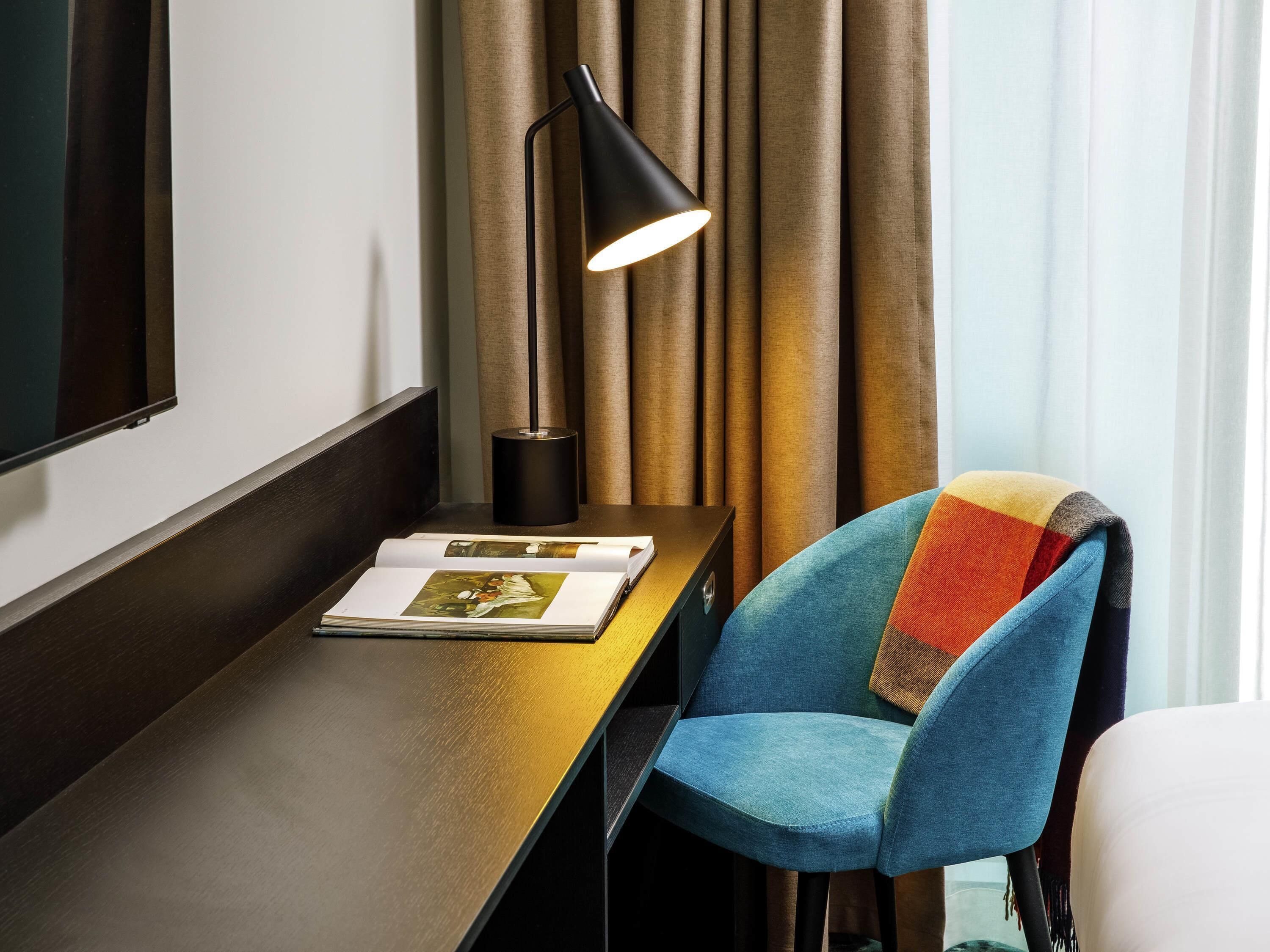 Quarto de hotel com uma escrivaninha, uma luminária de mesa preta, uma cadeira azul e uma janela com cortinas.