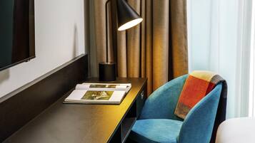 Quarto de hotel com uma escrivaninha, uma luminária de mesa preta, uma cadeira azul e uma janela com cortinas.
