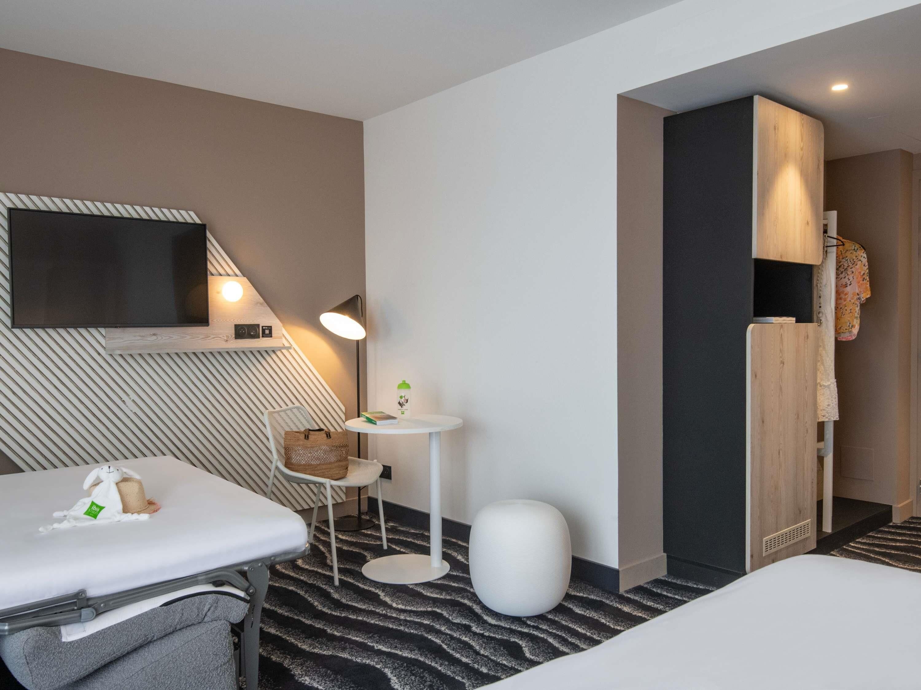 Quarto de hotel com cama, uma pequena mesa com cadeira, televisão na parede, abajur e guarda-roupa.