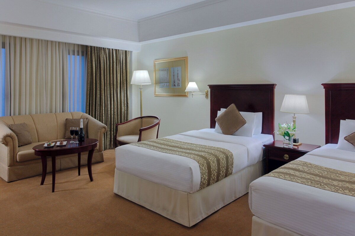 Quarto de hotel com duas camas, um sofá, uma mesinha e um abajur.