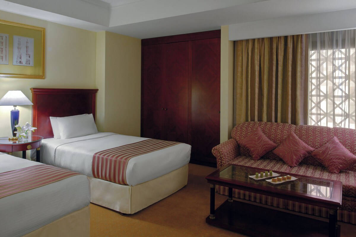 Quarto de hotel com duas camas, um sofá, uma mesa de centro, uma mesinha de cabeceira e um guarda-roupa espaçoso.