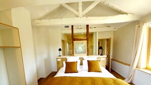 Quarto com uma cama grande, teto de madeira, banheiro visível por uma porta de vidro e janela com cortinas.