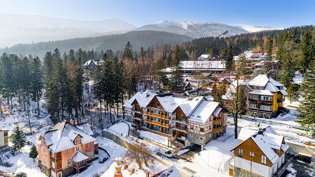 Estância de montanha coberta de neve com vários edifícios de madeira, incluindo um grande chalé com um teleférico visível ao fundo.