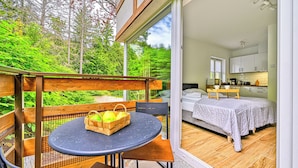 Uma varanda com mesa e cadeiras, uma cama com colcha branca e uma área de cozinha visível através de uma porta aberta.