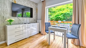 Uma sala de estar moderna com televisão de tela plana fixada em uma parede texturizada, um armário branco com gavetas, uma mesa de jantar com cadeiras e uma grande janela que oferece vista para o verde.