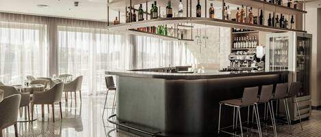 Uma área de bar moderna com balcão curvo, várias prateleiras cheias de garrafas de bebidas e um espaço com cadeiras e mesas.