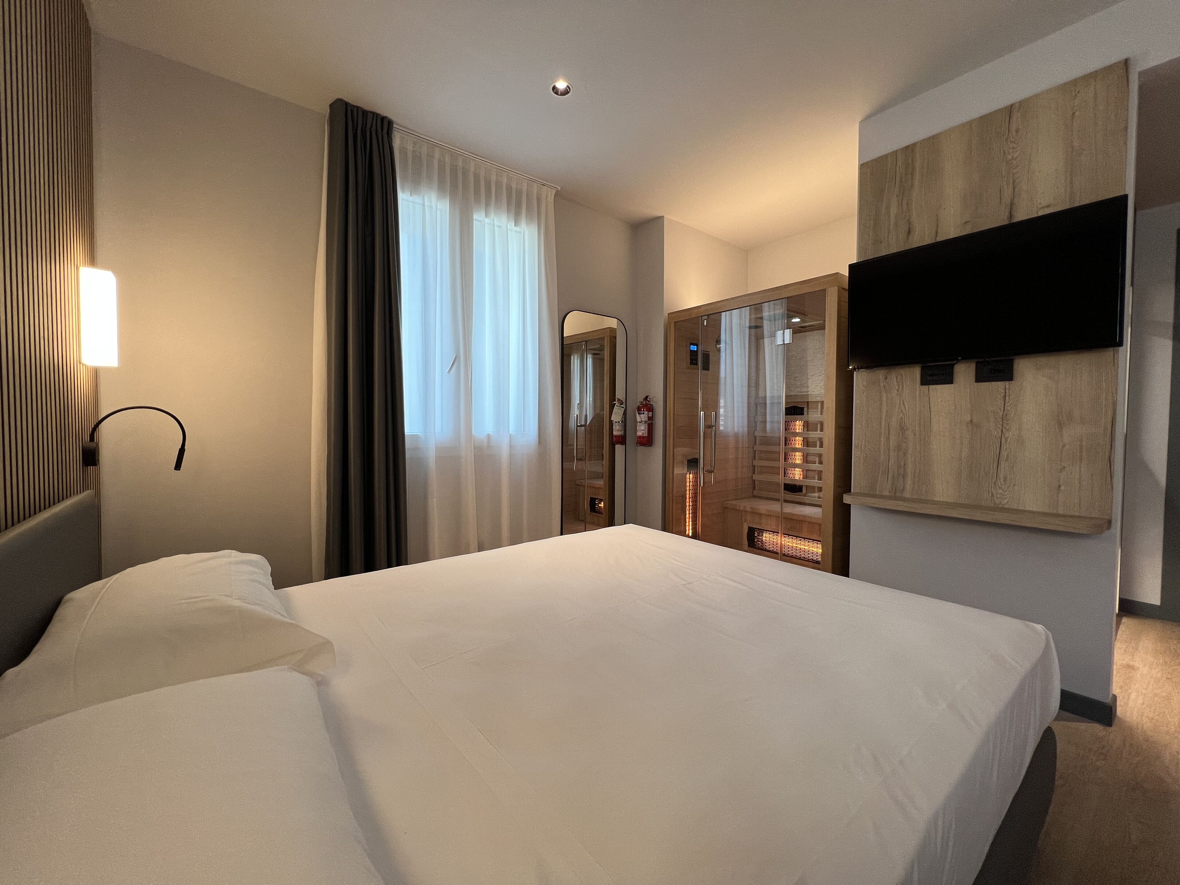 Quarto de hotel com uma cama grande, luminária, espelho, guarda-roupa e televisão fixada na parede.