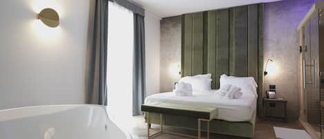 Quarto de hotel moderno com uma banheira grande, cama arrumada com toalhas, mesa de cabeceira com abajur e vista para a cidade pela janela.