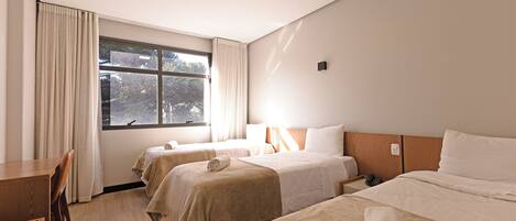 Quarto de hotel com duas camas, uma escrivaninha, uma cadeira, janela com cortinas e mesa de cabeceira com abajur.