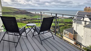 Varanda com duas cadeiras e uma mesa, com vista para a paisagem costeira com colinas e uma praia.