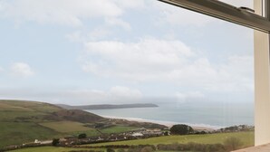 Vista da janela sobre uma paisagem costeira com colinas ondulantes, uma praia e uma pequena aldeia.
