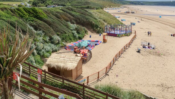 Uma praia com uma área cercada, contendo um parquinho e um edifício com telhado de palha.