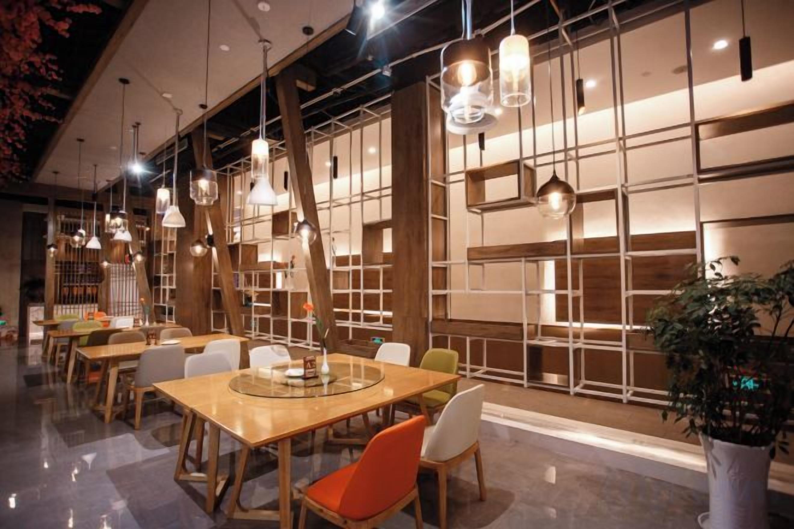 Interior de restaurante com mesas e cadeiras de madeira, luminárias pendentes e uma grande planta em vaso.