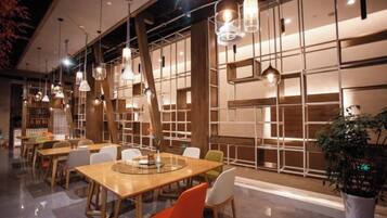 Interior de restaurante com mesas e cadeiras de madeira, luminárias pendentes e uma grande planta em vaso.