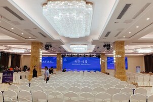 Uma sala de conferências com fileiras de cadeiras brancas, um palco com fundo azul e um lustre grande.