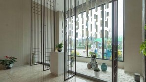Quarto de hotel moderno com uma grande porta de vidro deslizante que dá para uma varanda. O quarto possui piso de azulejo, um balcão de recepção branco e algumas plantas em vasos.