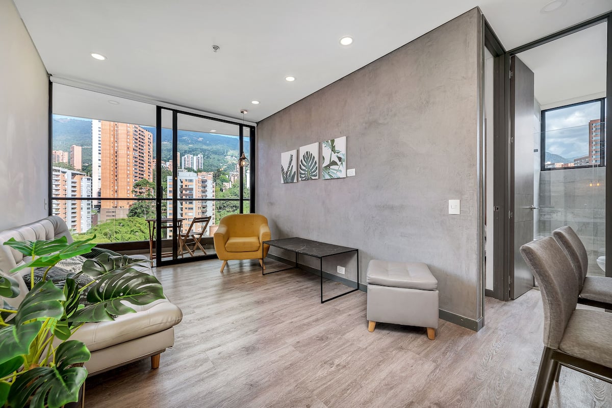 Uma sala de estar moderna com sofá, poltrona, apoio para os pés, uma mesa pequena e uma janela grande com vista para a cidade.