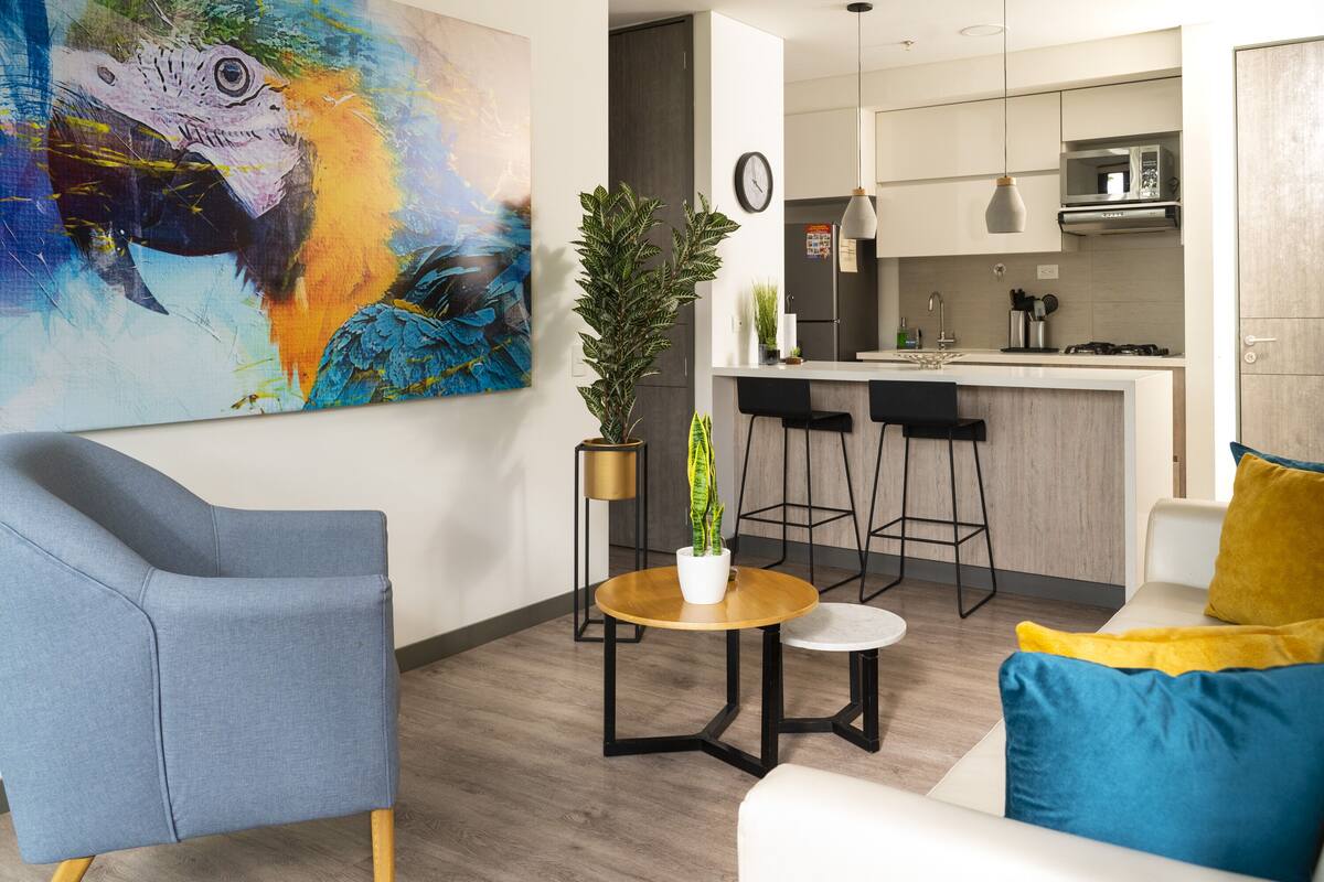 Uma sala de estar moderna com uma poltrona azul, uma mesa de centro redonda de madeira e um sofá branco com almofadas amarelas e azuis. Um grande quadro abstrato está na parede, e há uma planta em vaso sobre a mesa de centro.