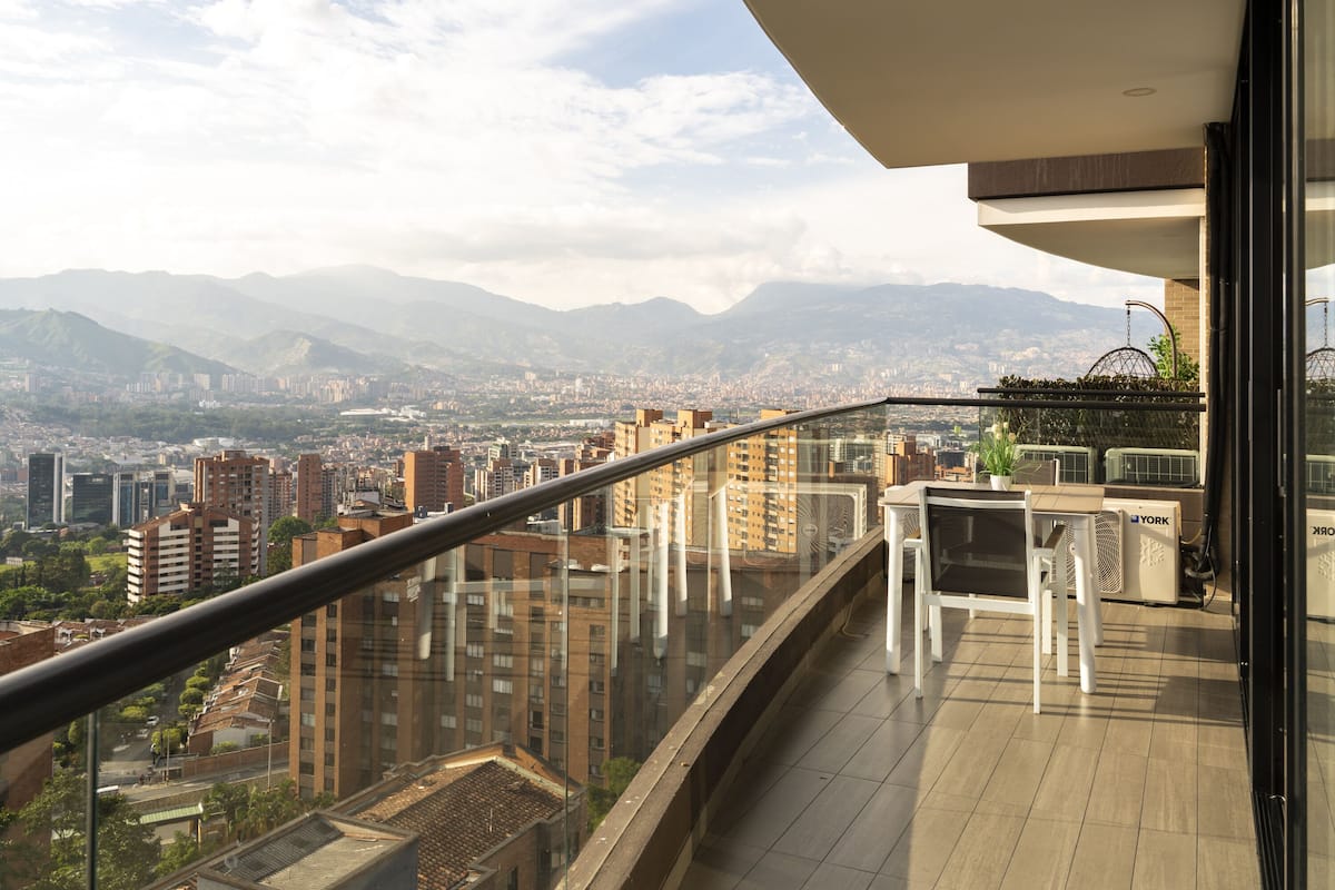 Varanda com guarda-corpo de vidro, vista para a cidade e montanhas.