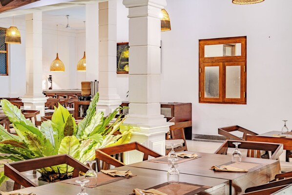 Interior de restaurante com mesas e cadeiras de madeira, luminárias pendentes e plantas verdes grandes.